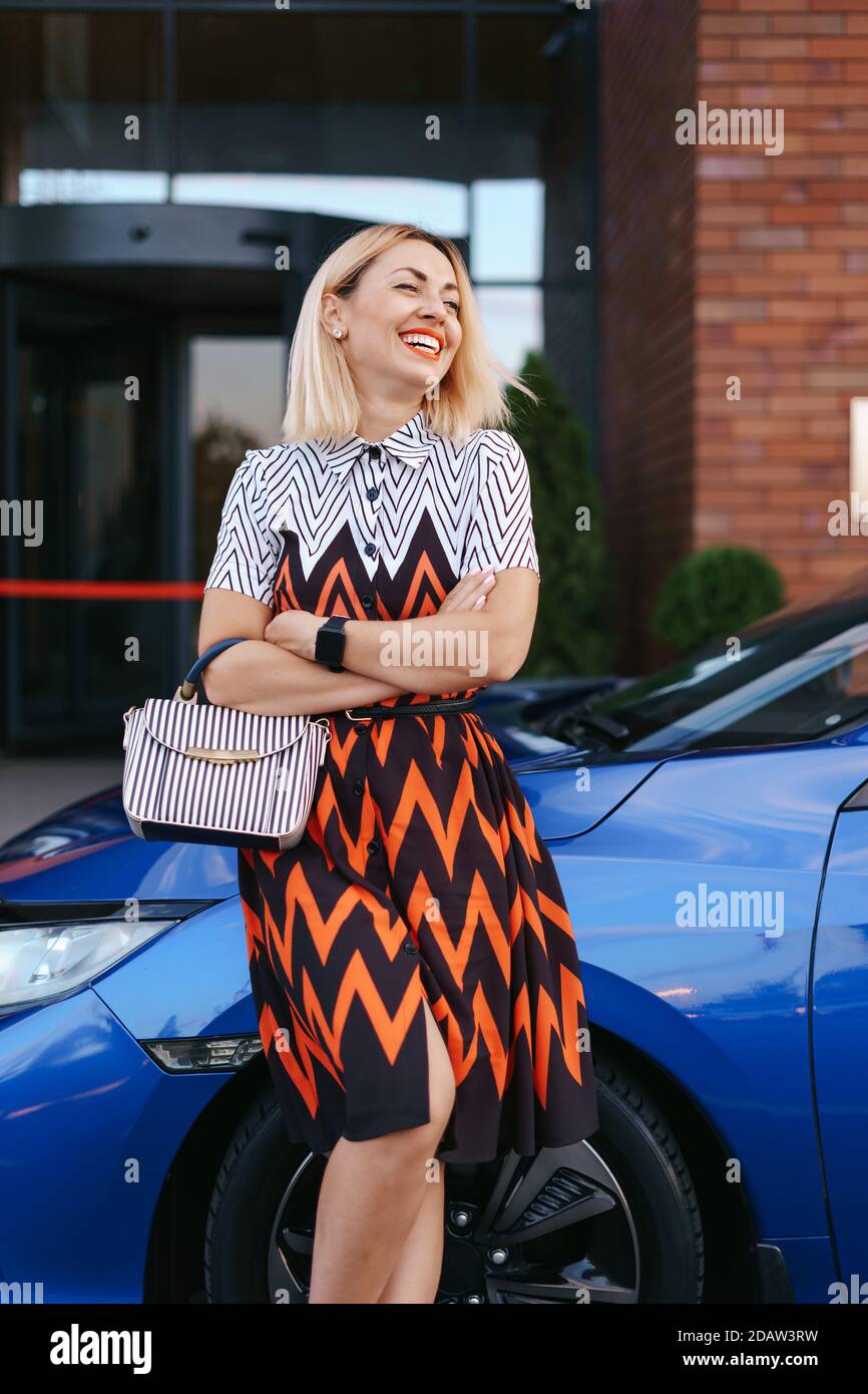 Stunning young woman waring dress posing in front of her car outdoors ...