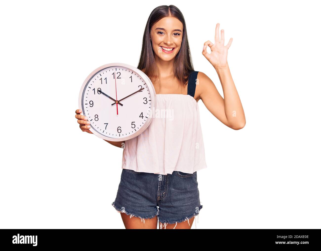 Young hispanic woman holding big clock doing ok sign with fingers ...