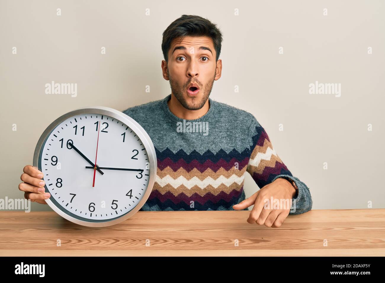 Young handsome man holding big clock scared and amazed with open mouth ...