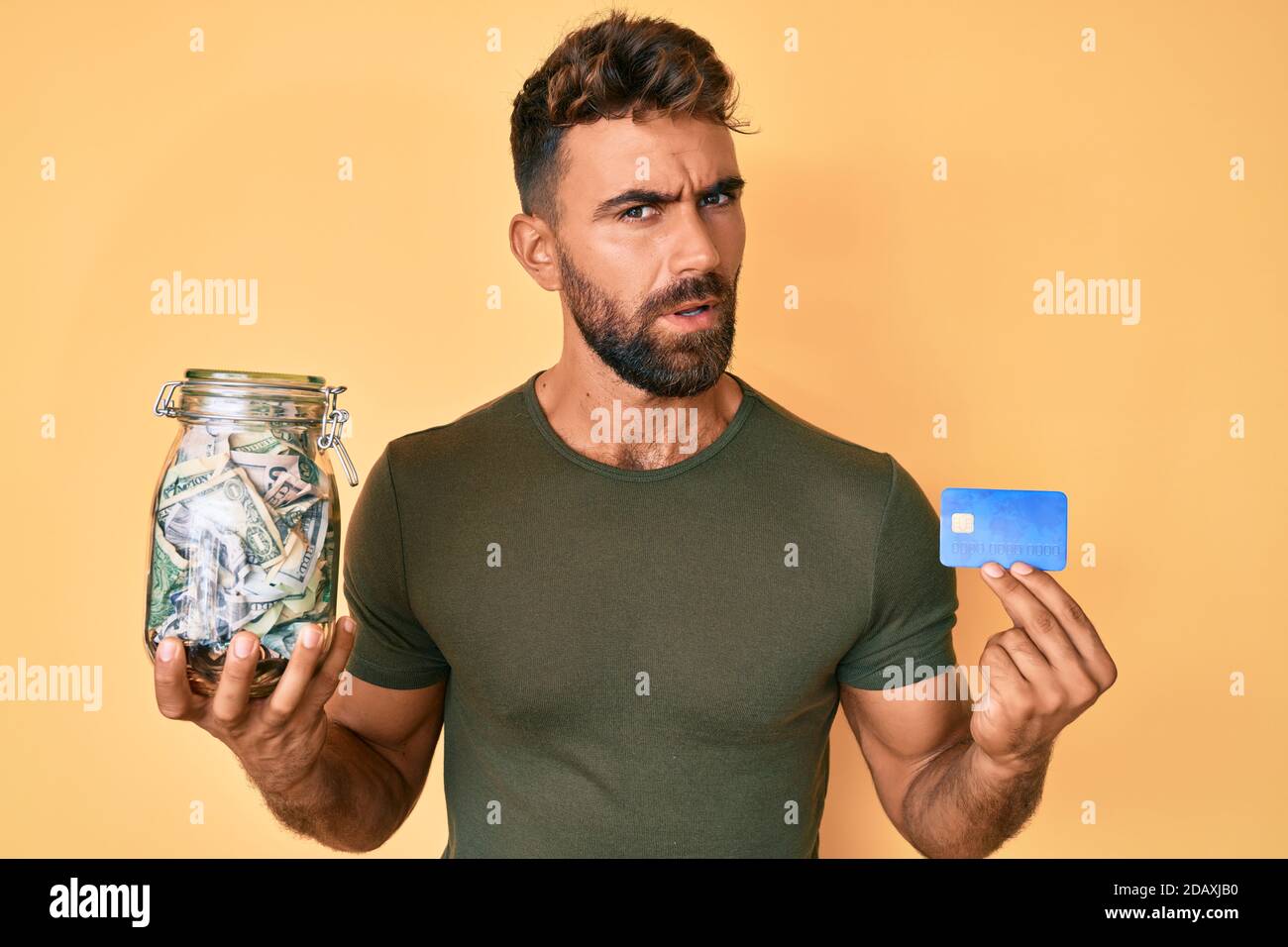 Young hispanic man holding credit car and jar with dollars in shock ...
