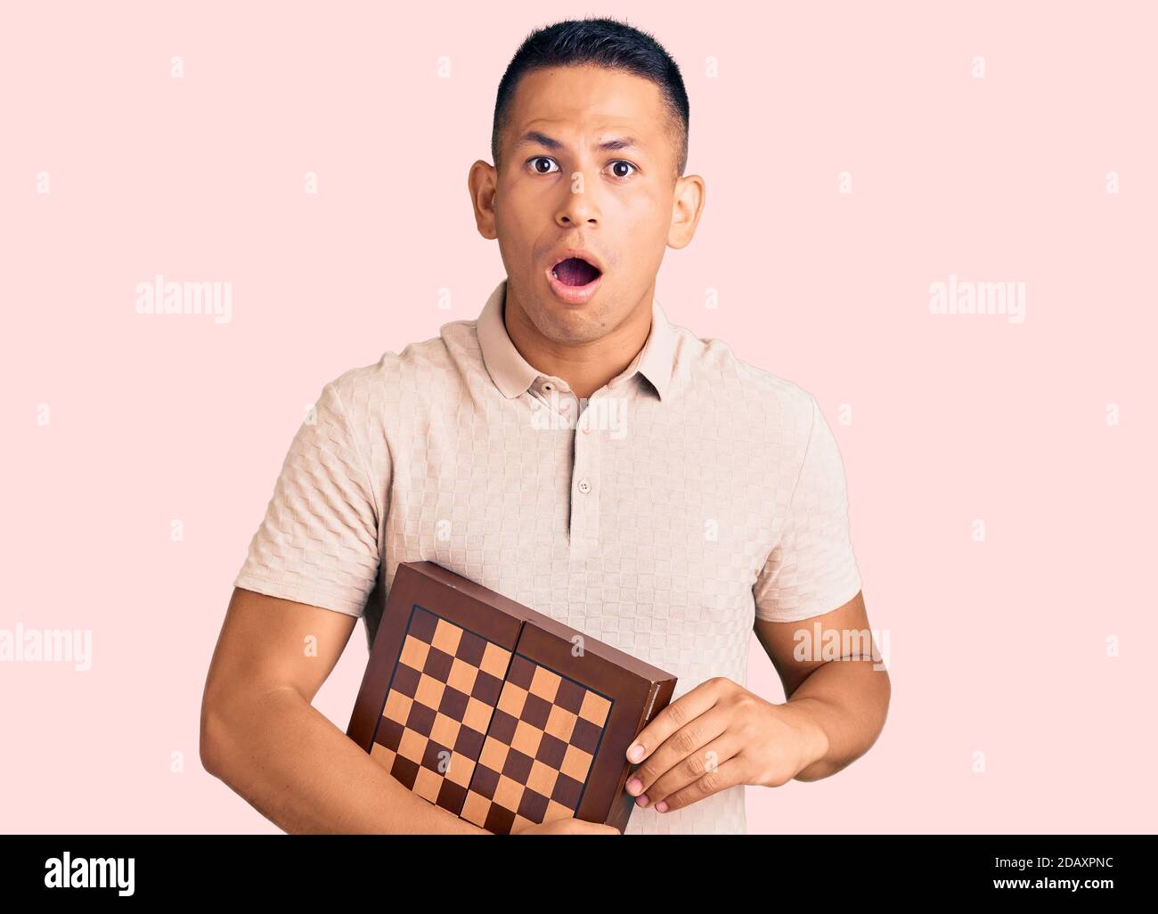 Young handsome latin man holding chess scared and amazed with open ...