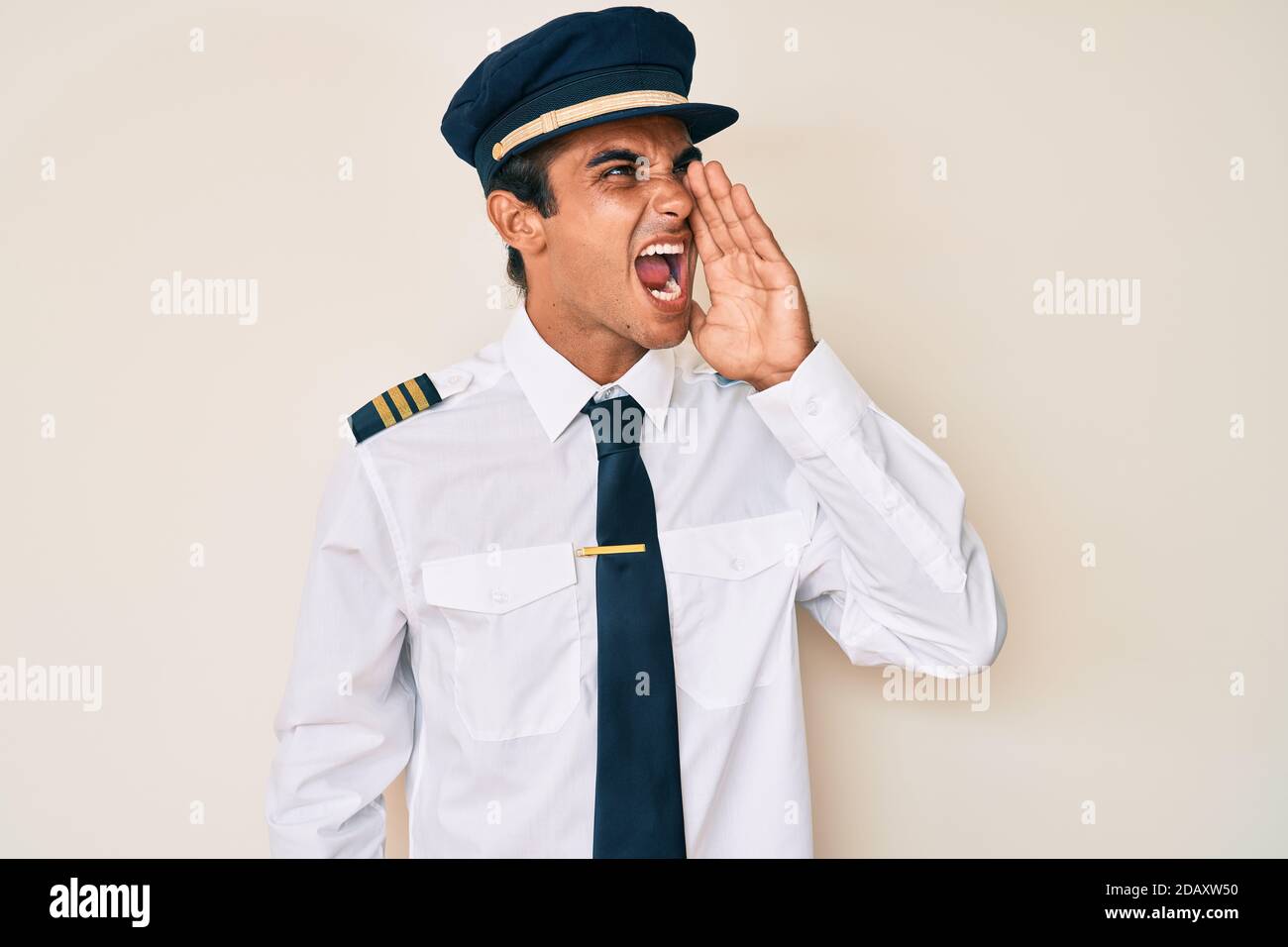 Young hispanic man wearing airplane pilot uniform shouting and ...