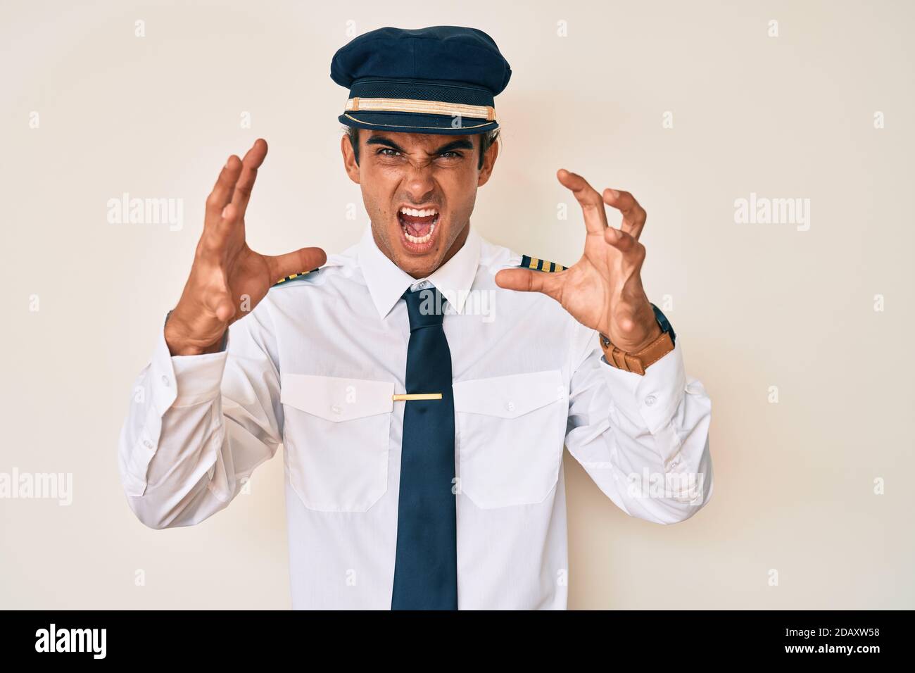 Young hispanic man wearing airplane pilot uniform shouting frustrated ...