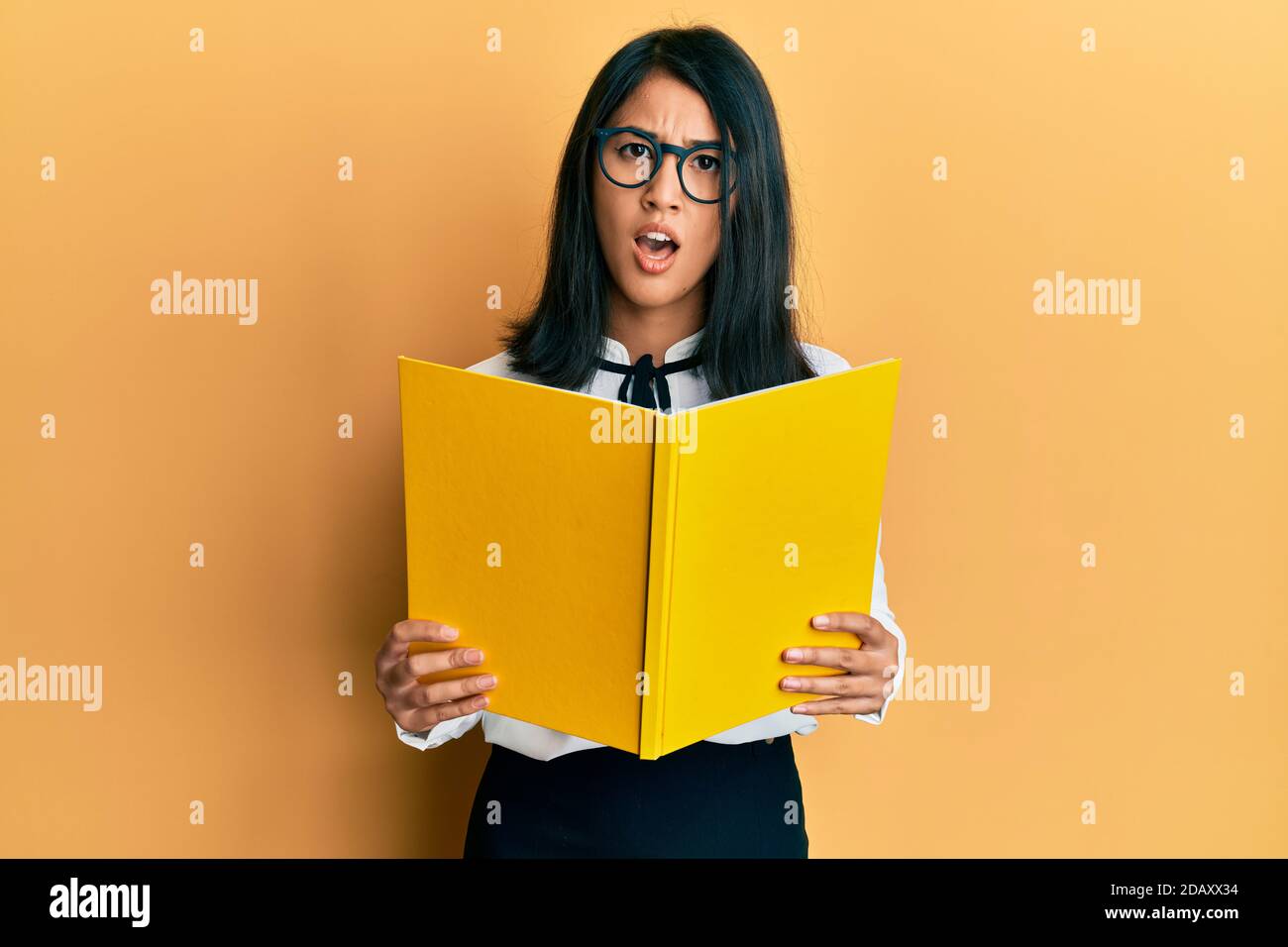Beautiful asian young woman reading a book wearing glasses in shock ...