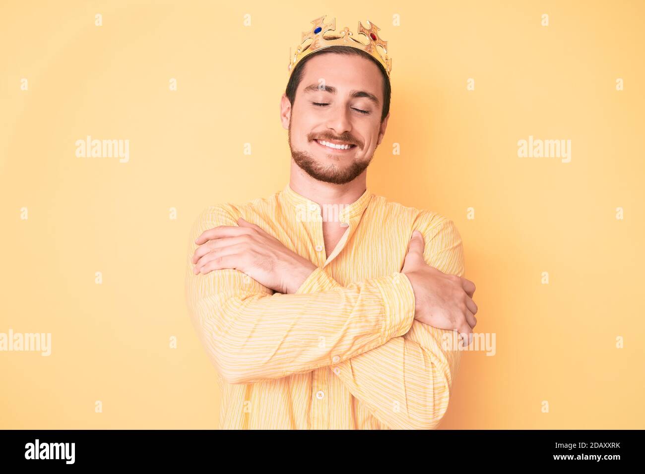 Young handsome man wearing king crown hugging oneself happy and ...