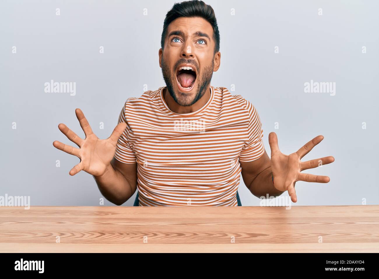 Handsome hispanic man wearing casual clothes sitting on the table crazy ...