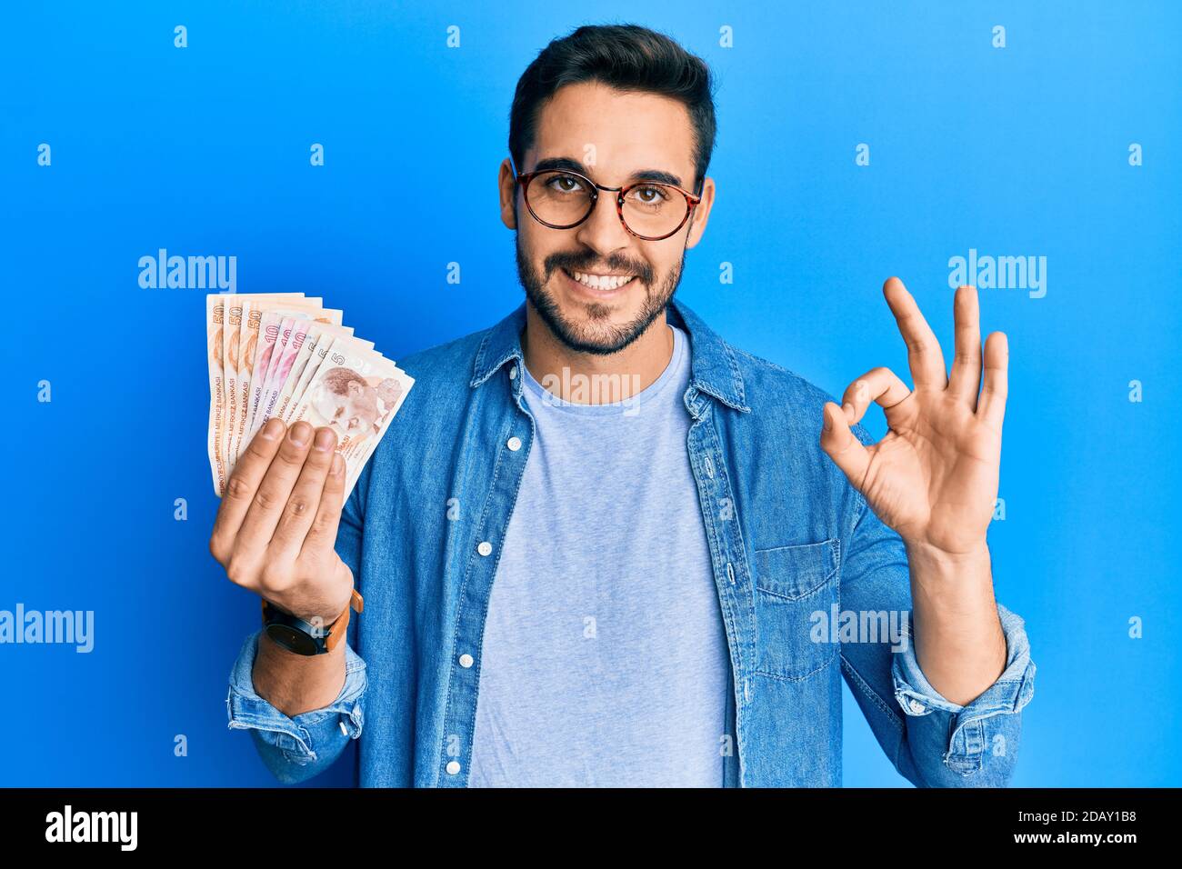 Young hispanic man holding turkish lira banknotes doing ok sign with ...