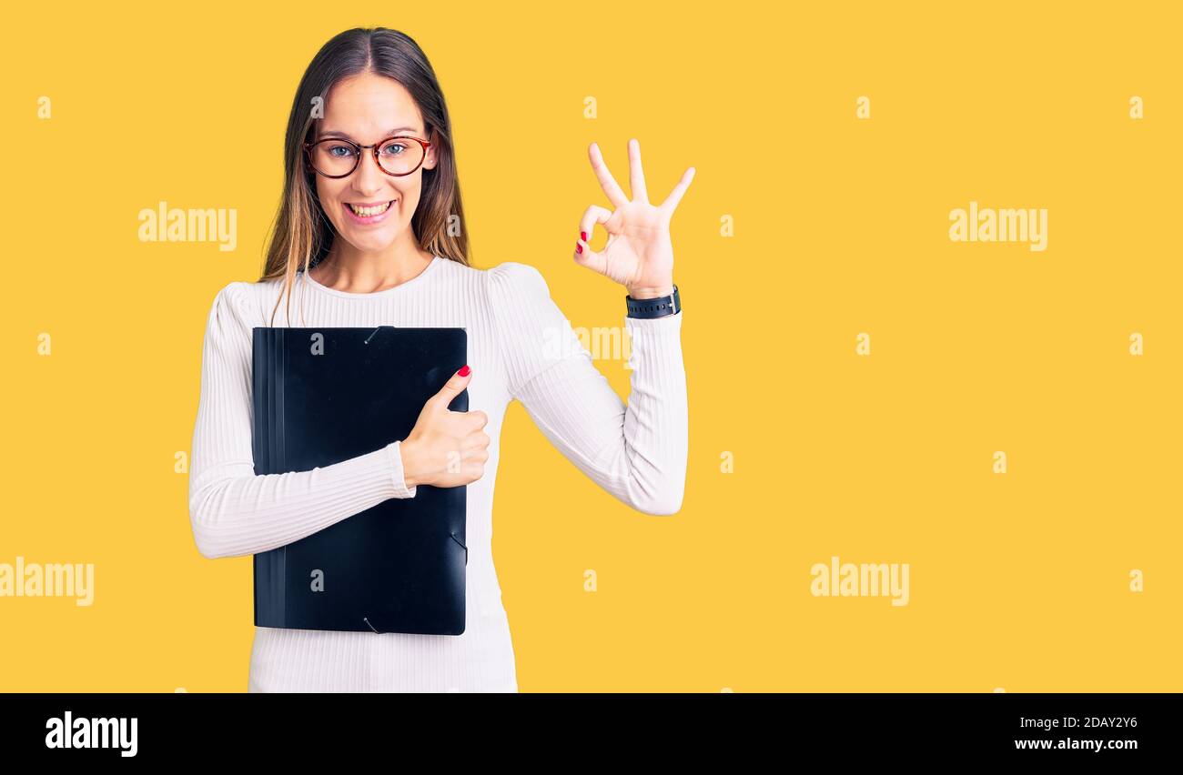 Beautiful brunette young woman holding business folder doing ok sign ...
