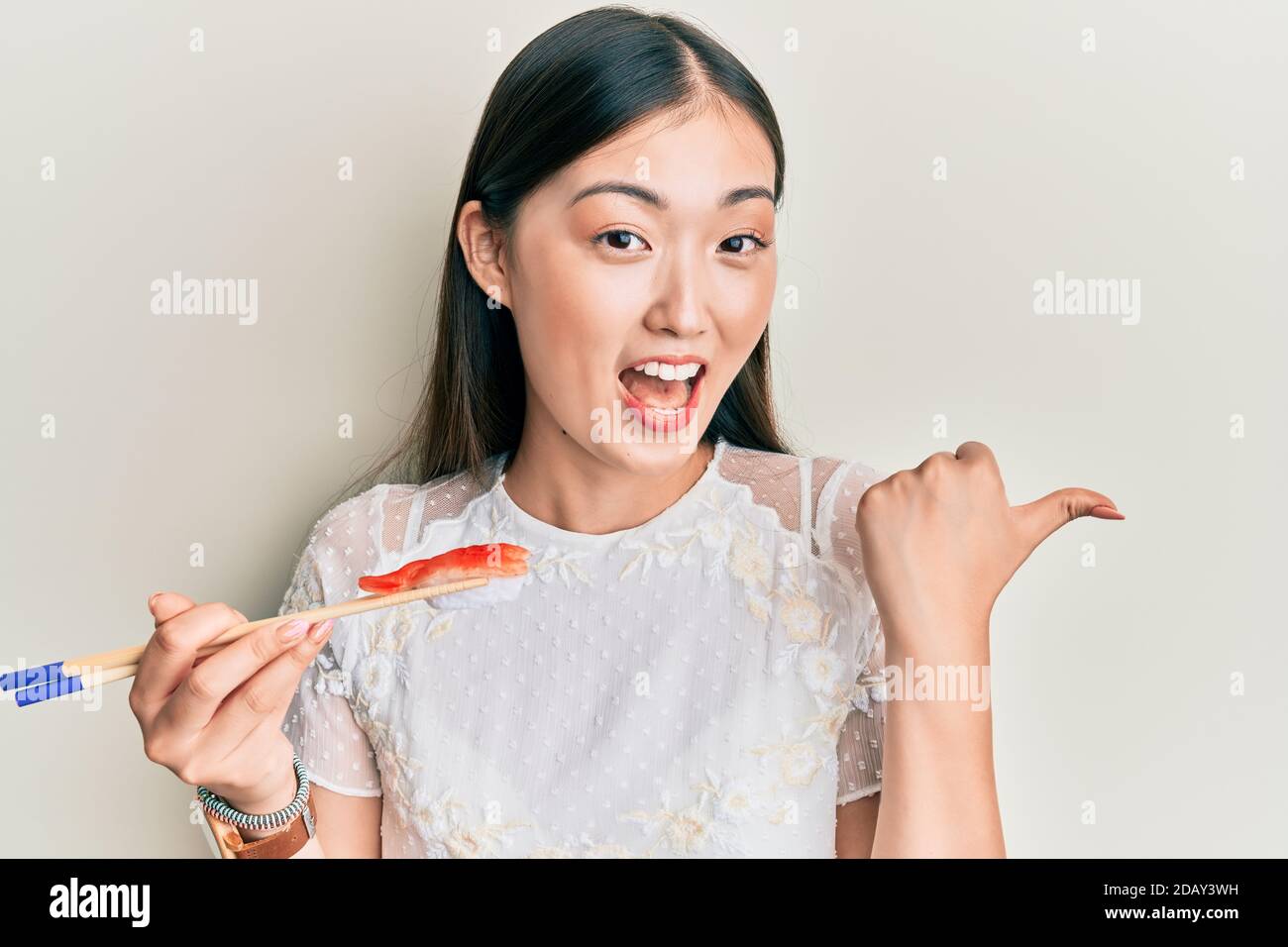 Young chinese woman eating prawn sushi using chopsticks pointing thumb ...
