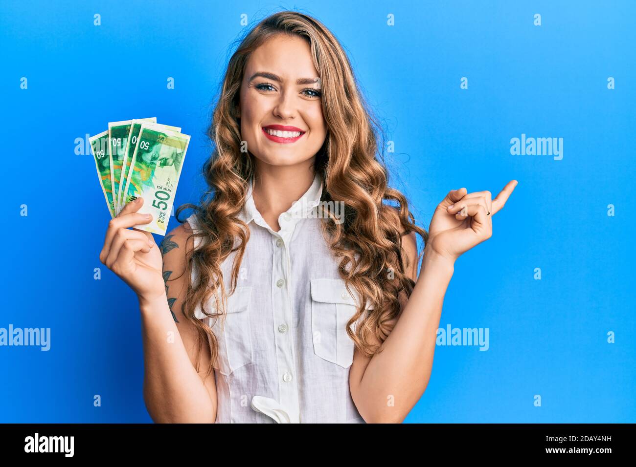 Young blonde girl holding israel shekels smiling happy pointing with ...