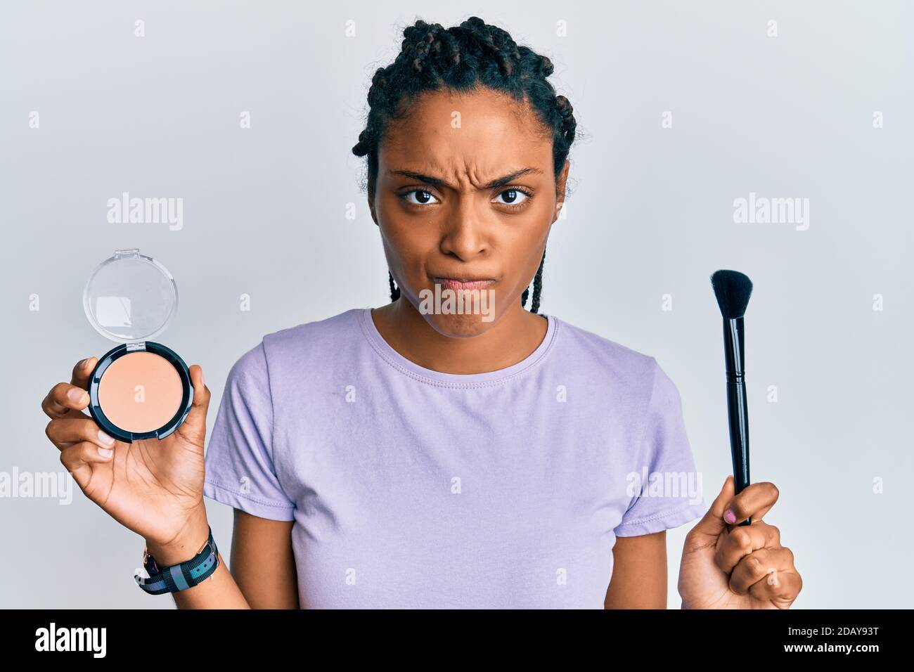 African american woman holding makeup brush and blush skeptic and ...