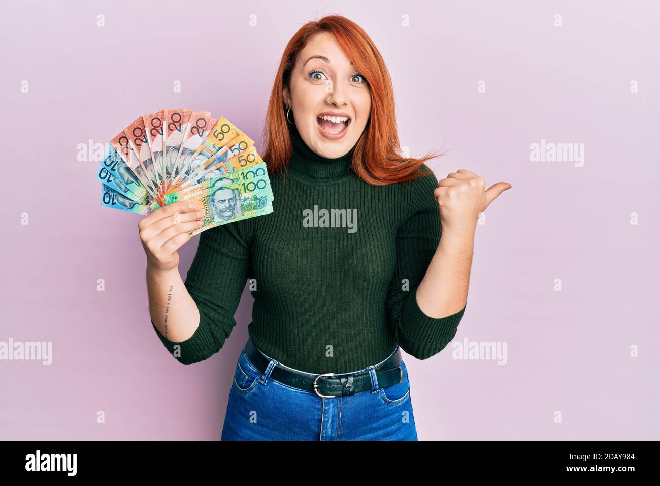 Beautiful Redhead Woman Holding Australian Dollars Pointing Thumb Up To The Side Smiling Happy 