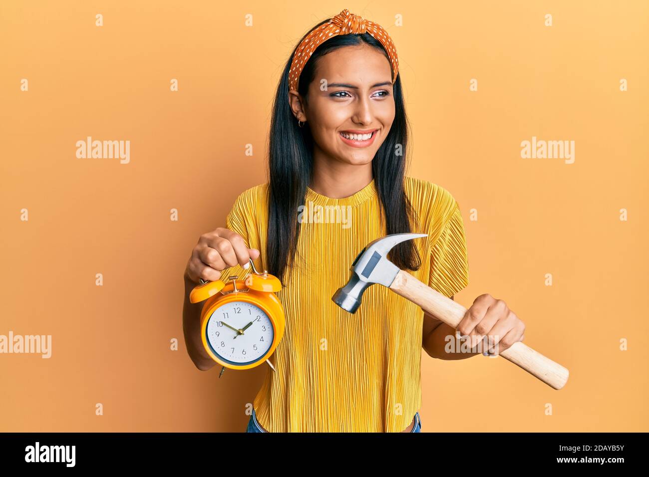 Young brunette woman holding alarm clock and hammer smiling looking to ...