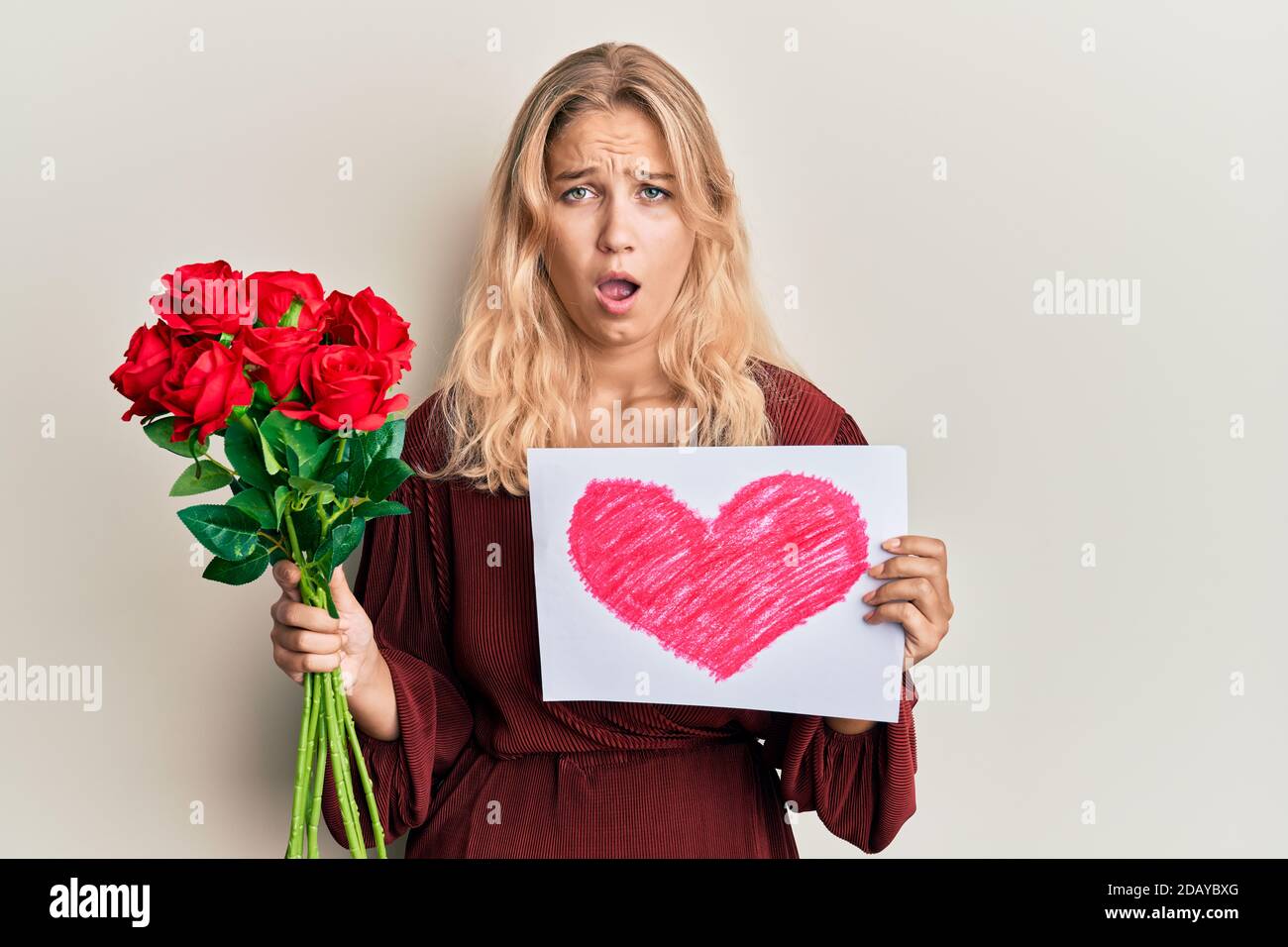 Young blonde girl holding heart draw and bouquet of roses afraid and ...