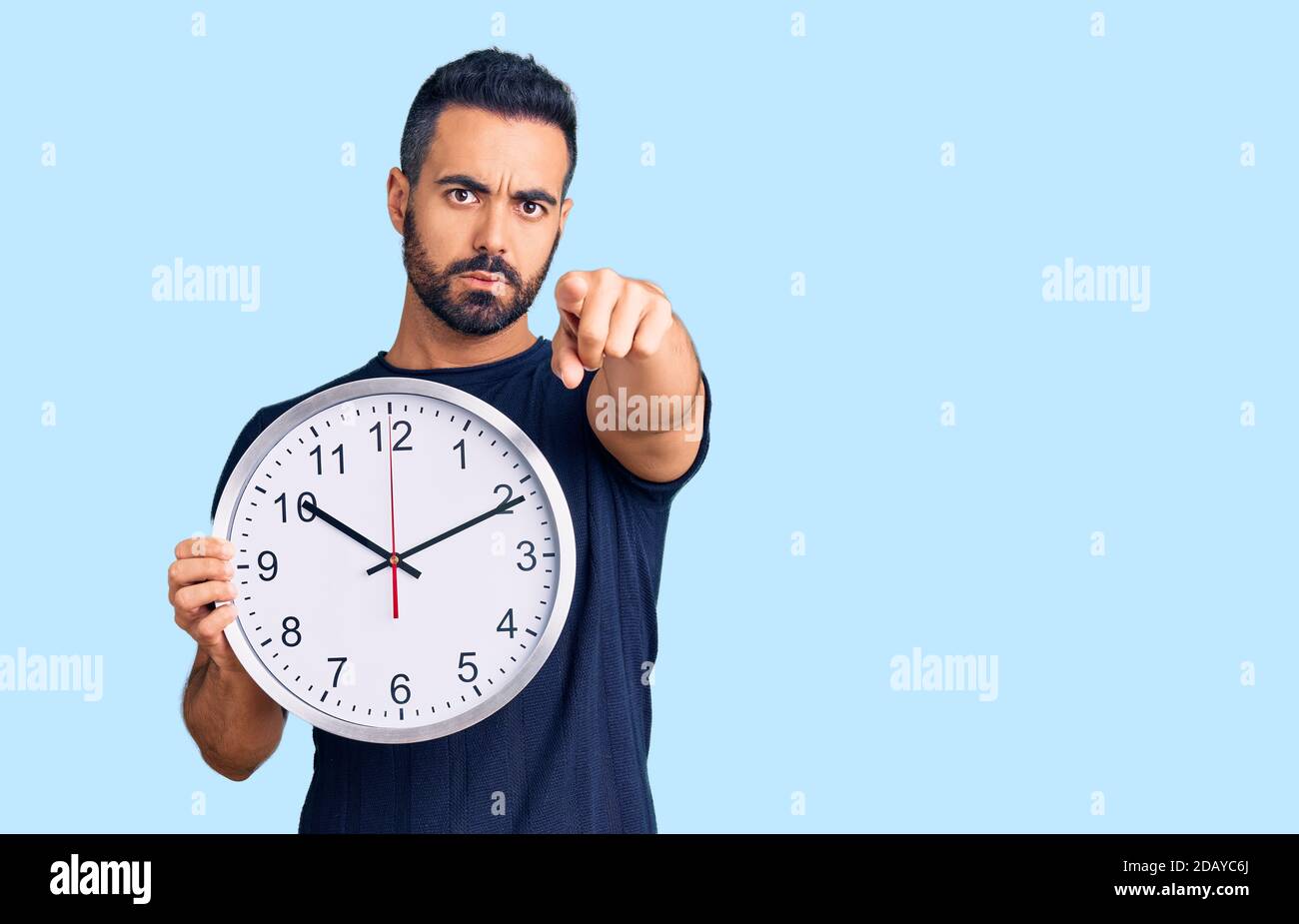 Young hispanic man holding big clock pointing with finger to the camera ...