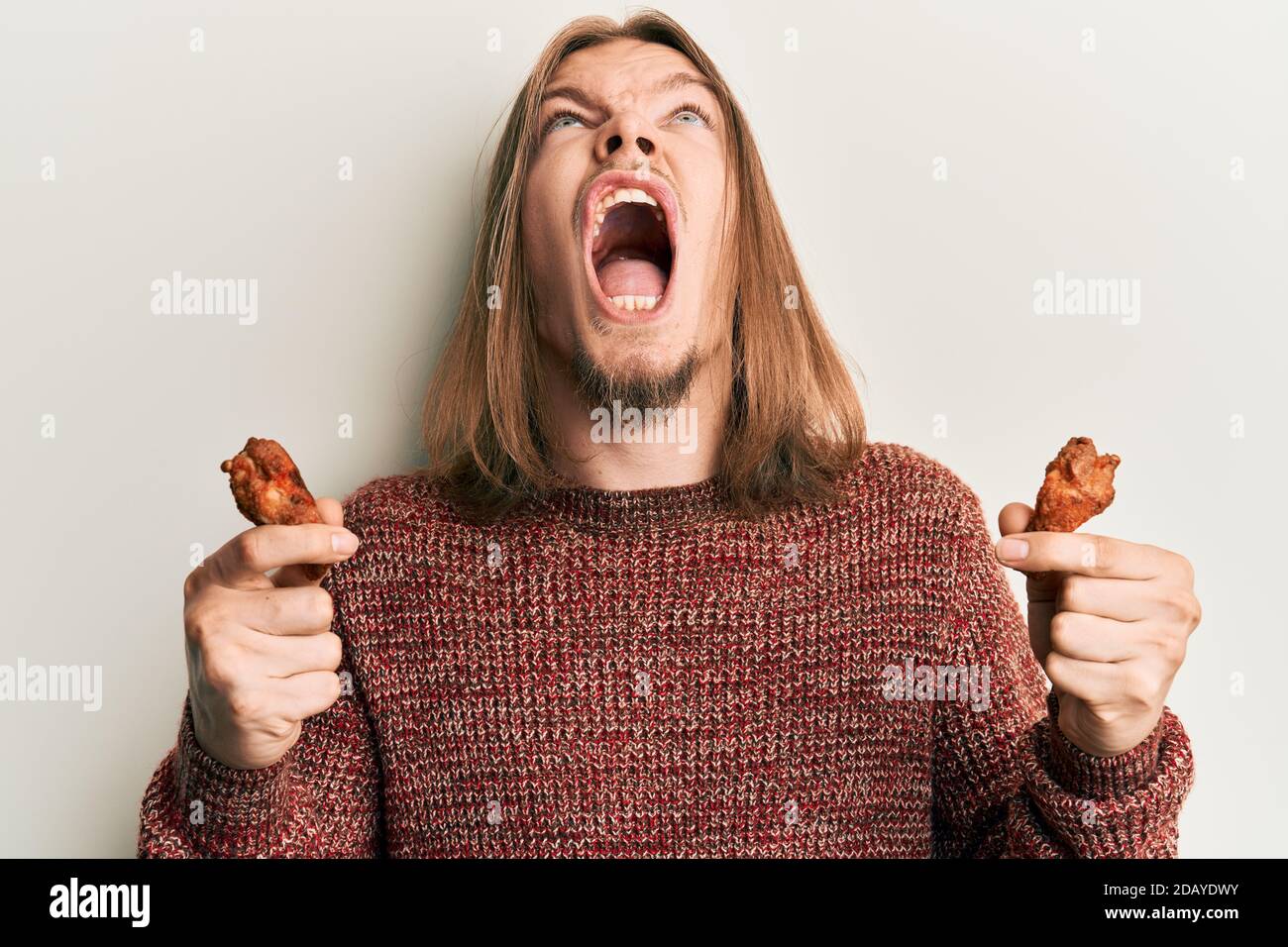 Handsome caucasian man with long hair eating chicken wings angry and ...
