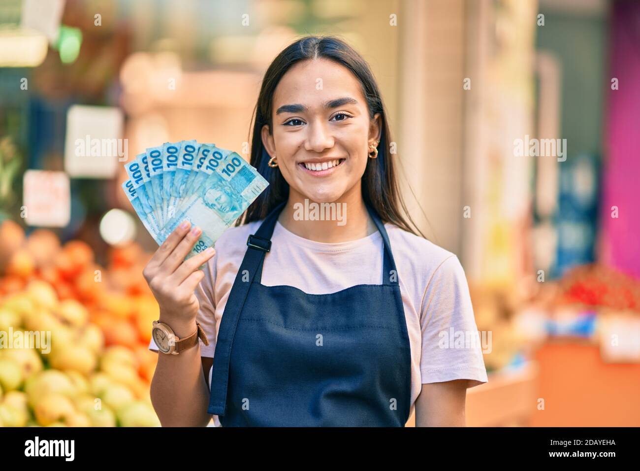 Young latin shopkeeper girl smiling happy holding brazil real banknotes ...