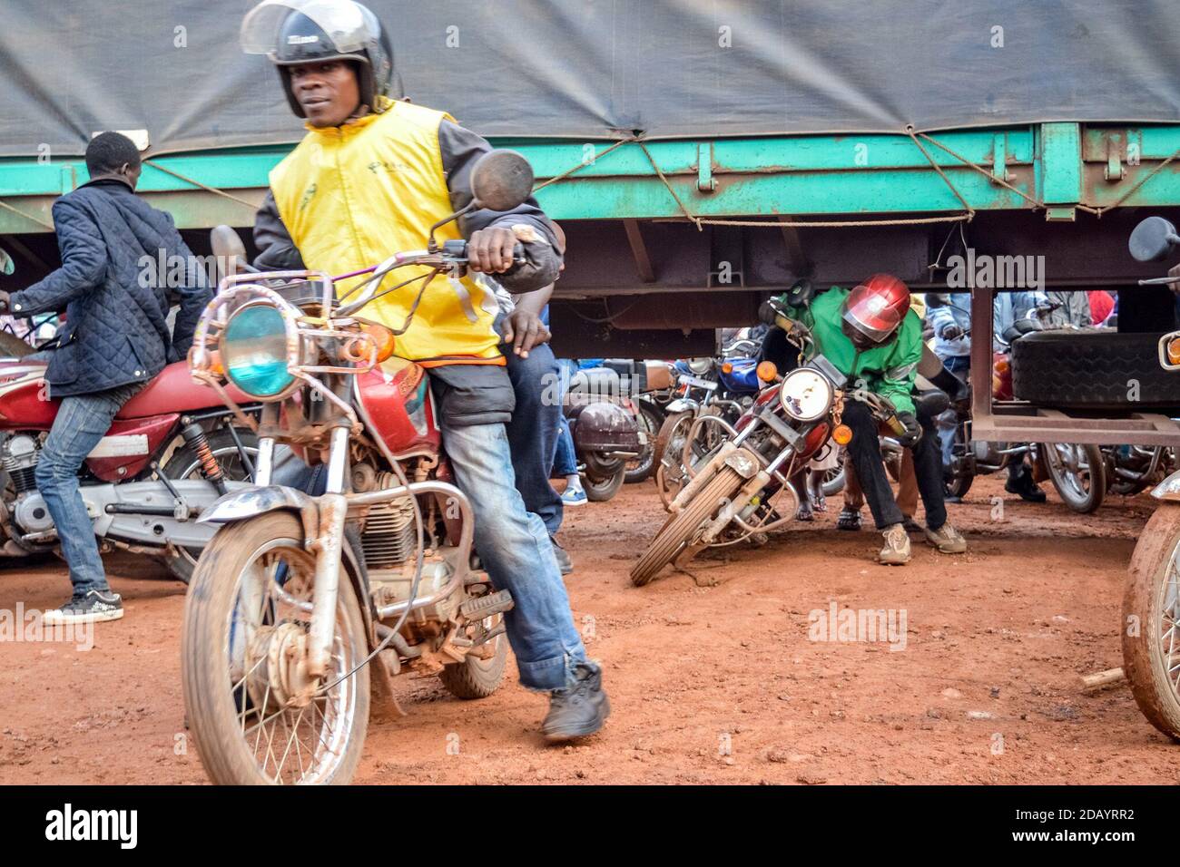 Boda boda motorcycle taxi operators pass under a stuck tractor trailer ...