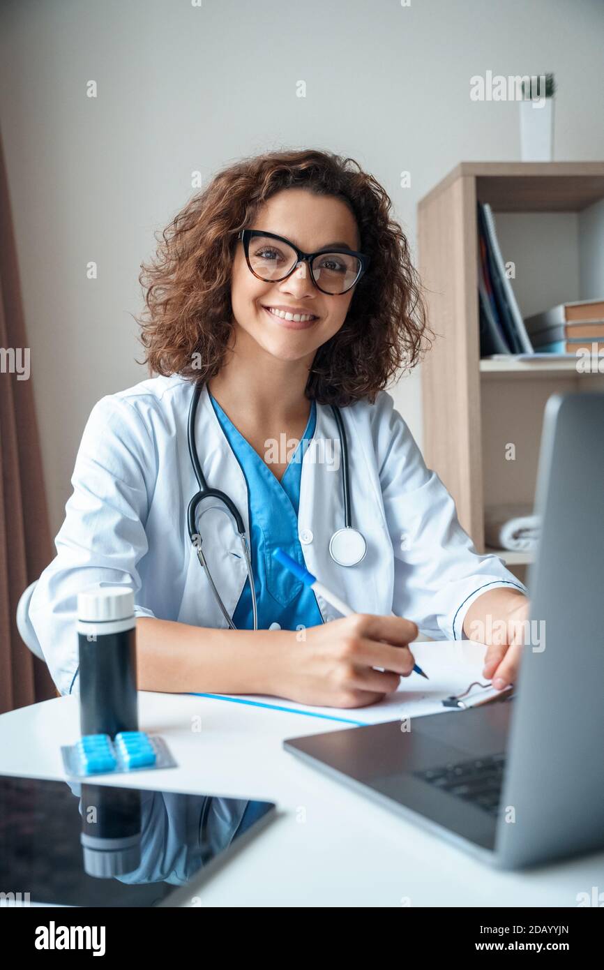 Vertical portrait of female doctor sitting on work desk and smiling at ...