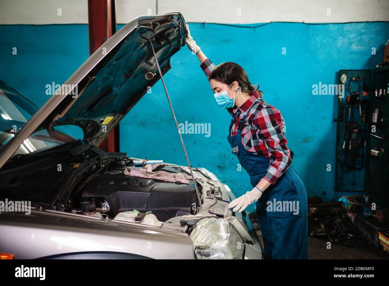 A young female mechanic in a uniform and medical mask opens the hood of ...