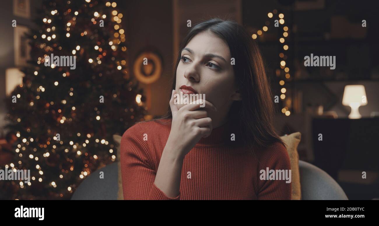 Lonely woman thinking with hand on chin at Christmas, tree with lights ...