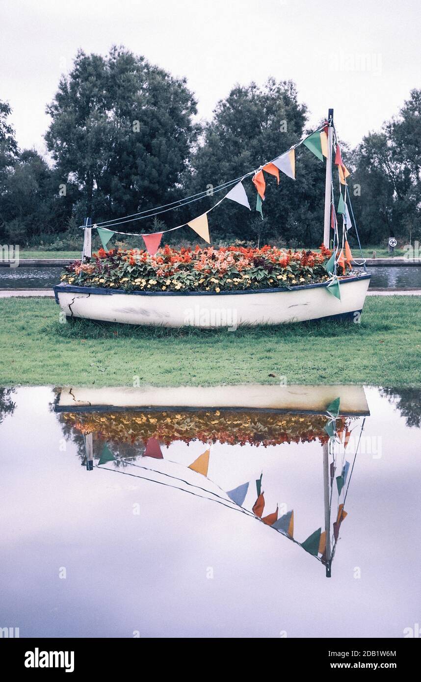 Reflection of a small rowing boat filled with flowers beside a river in ...