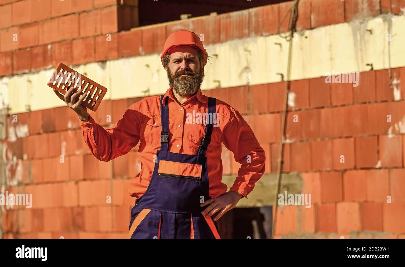 Builder hold brick. Bearded man wear hard hat at construction site. Brick building material used ...