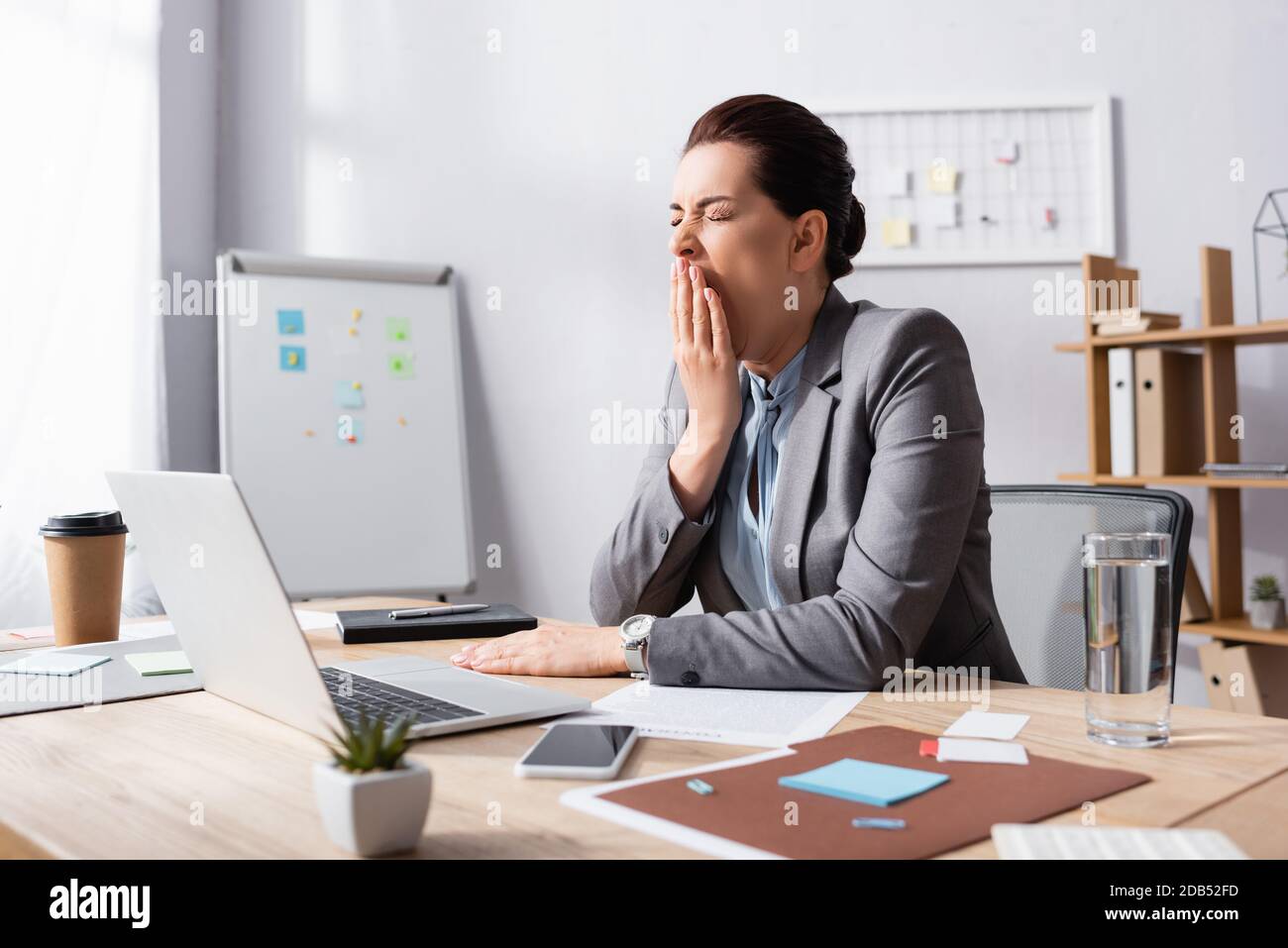 Sleepy businesswoman with hand near mouth yawning while sitting at ...