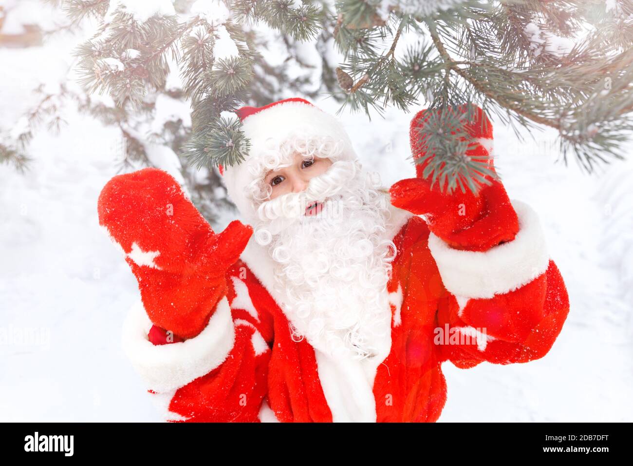 A child in a red santa claus costume in a winter forest shows hands in ...