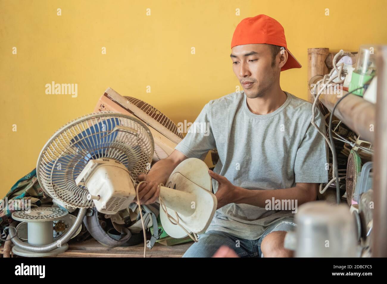 Asian electronics repairman holding fan while sitting around broken ...
