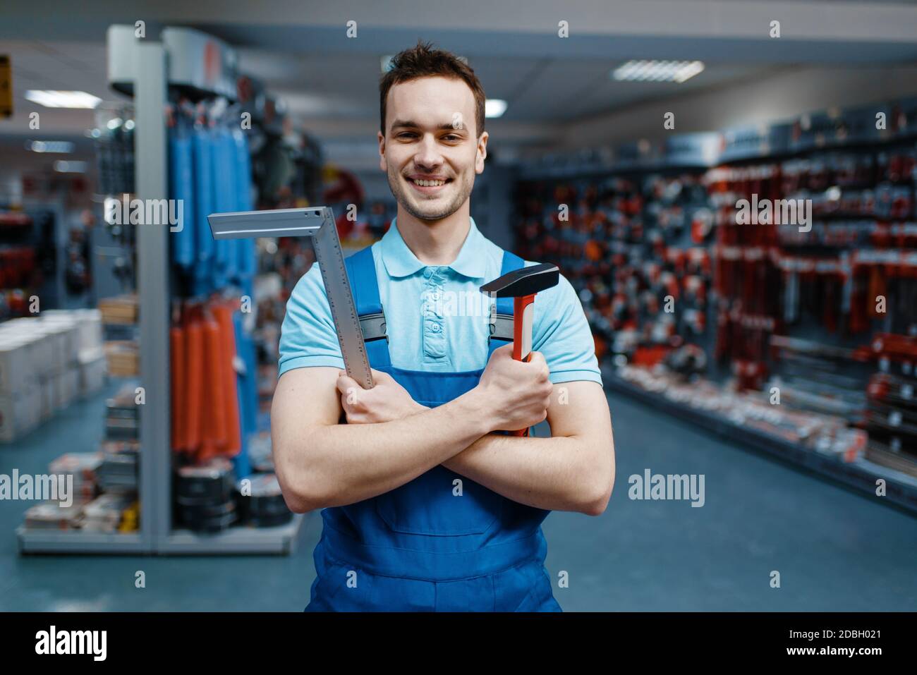 Smiling male worker in uniform holds hammer and corner in tool store ...