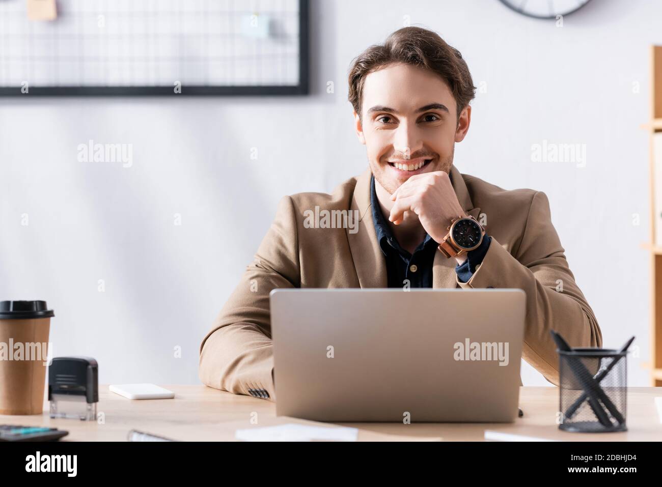 Smiling office worker looking at camera while sitting near laptop at ...
