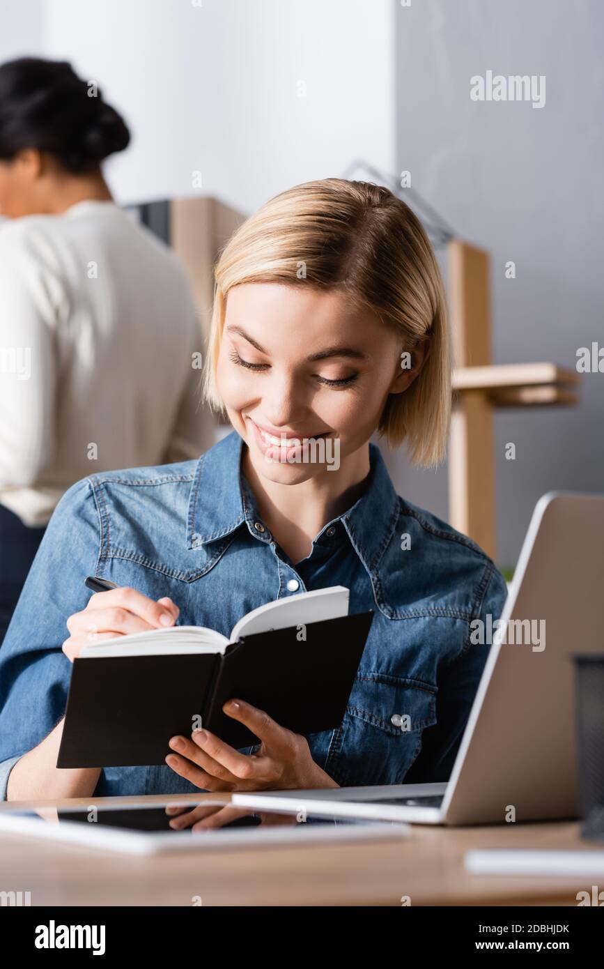 Happy blonde woman writing on notebook while sitting near laptop with ...