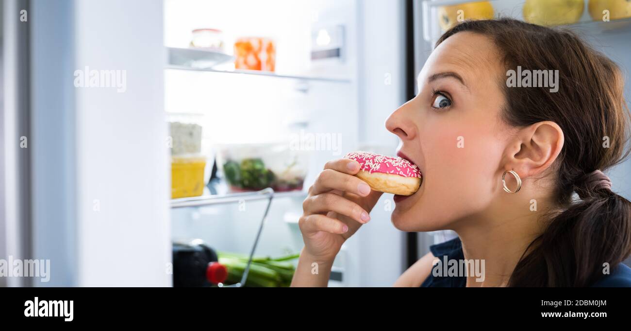 Hungry Young Woman Eating Donuts Near Fridge Stock Photo - Alamy