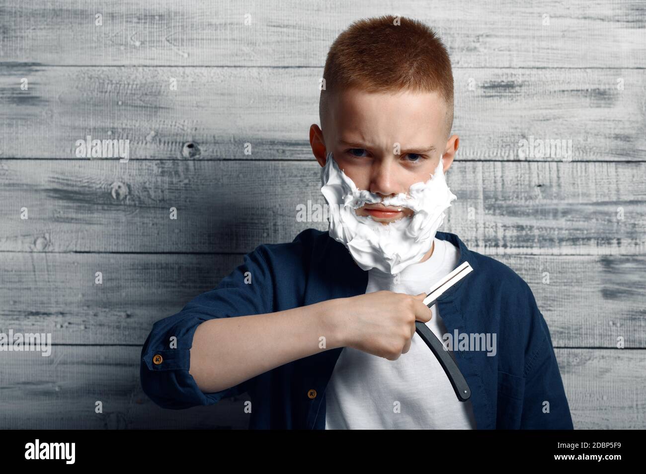 Serious little boy with shaving foam on his face holds a razor in ...