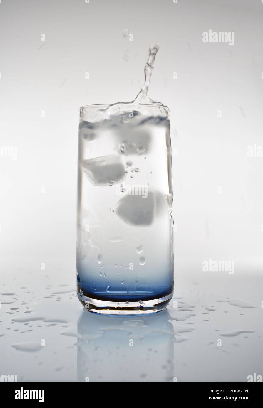 An ice cube dropping into a glass of water creating a splash. The glass has a blue colour at the ...