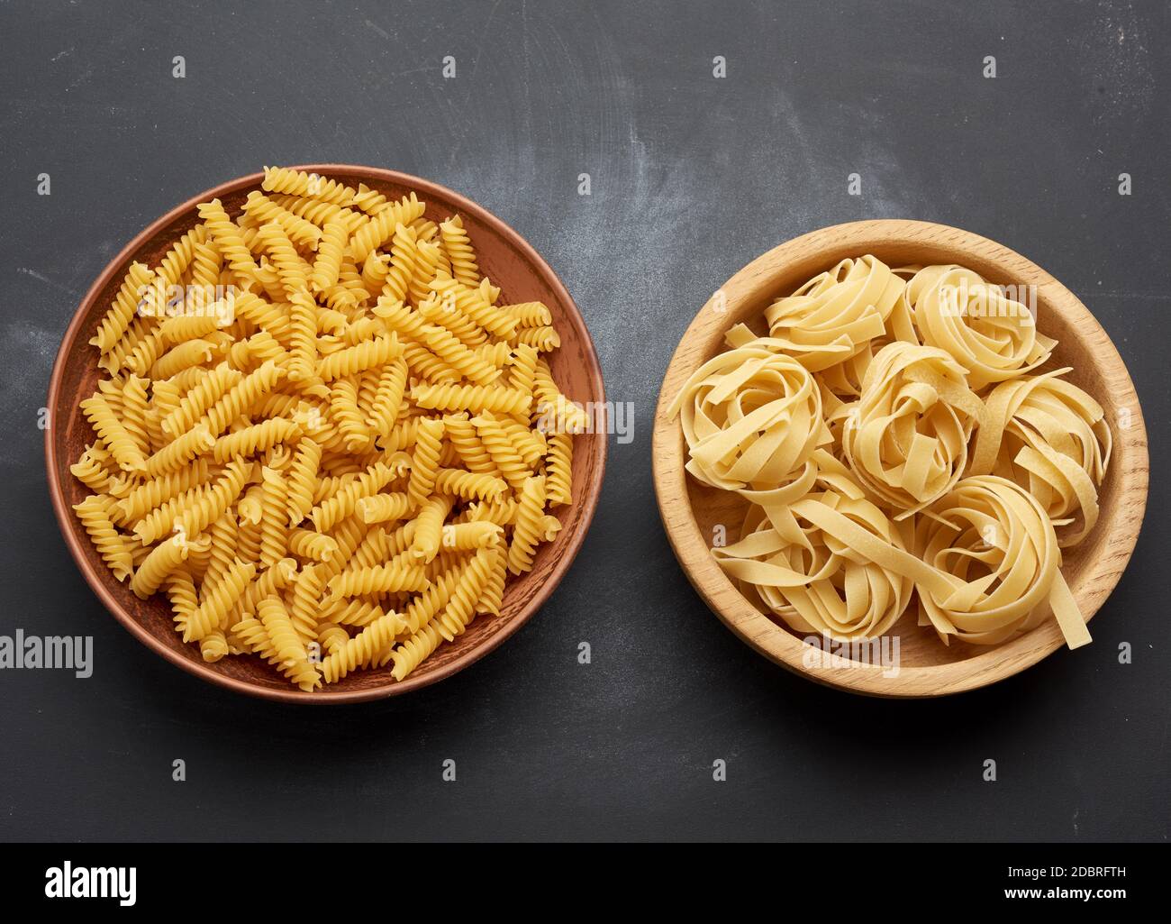 raw fusilli and fettuccine pasta in wooden plates on a black table, top ...
