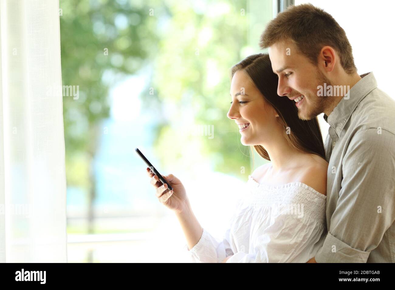 Side view of a couple checking smart phone standing next to window at ...