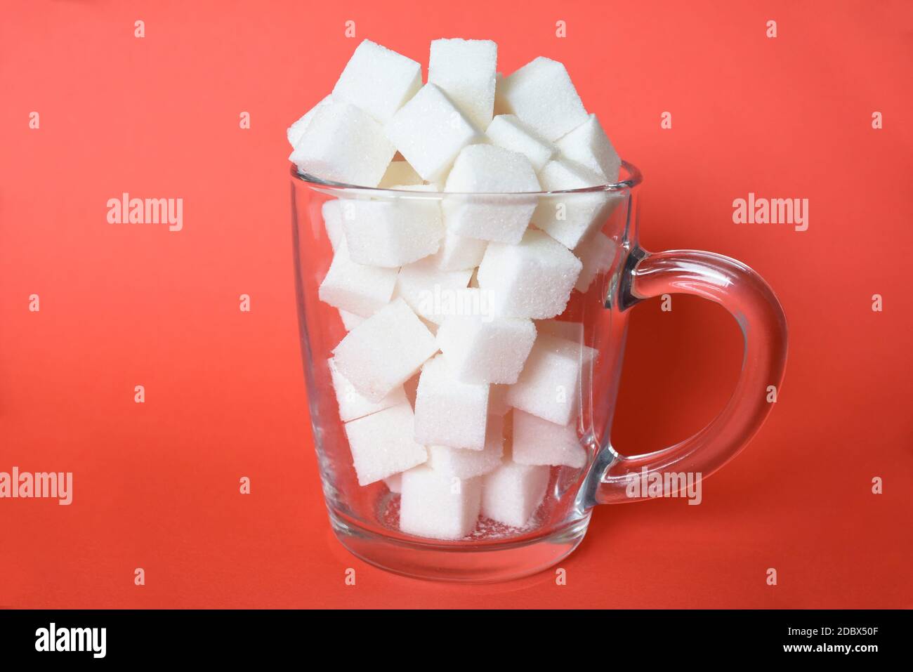 Transparent mug full of refined white sugar cubes on red background ...