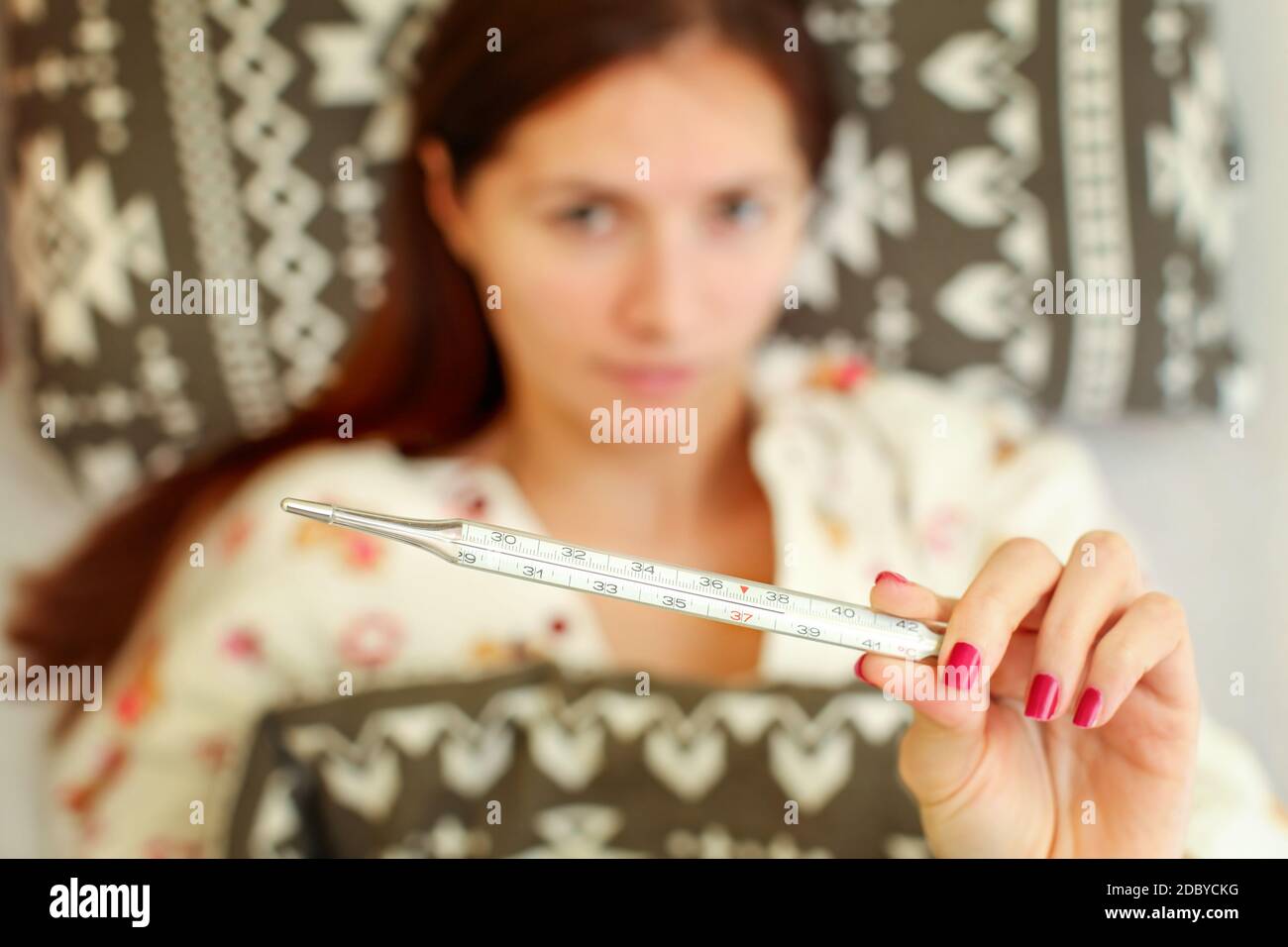 Sick young woman lying in the bed, covered with duvet, holding mercury ...