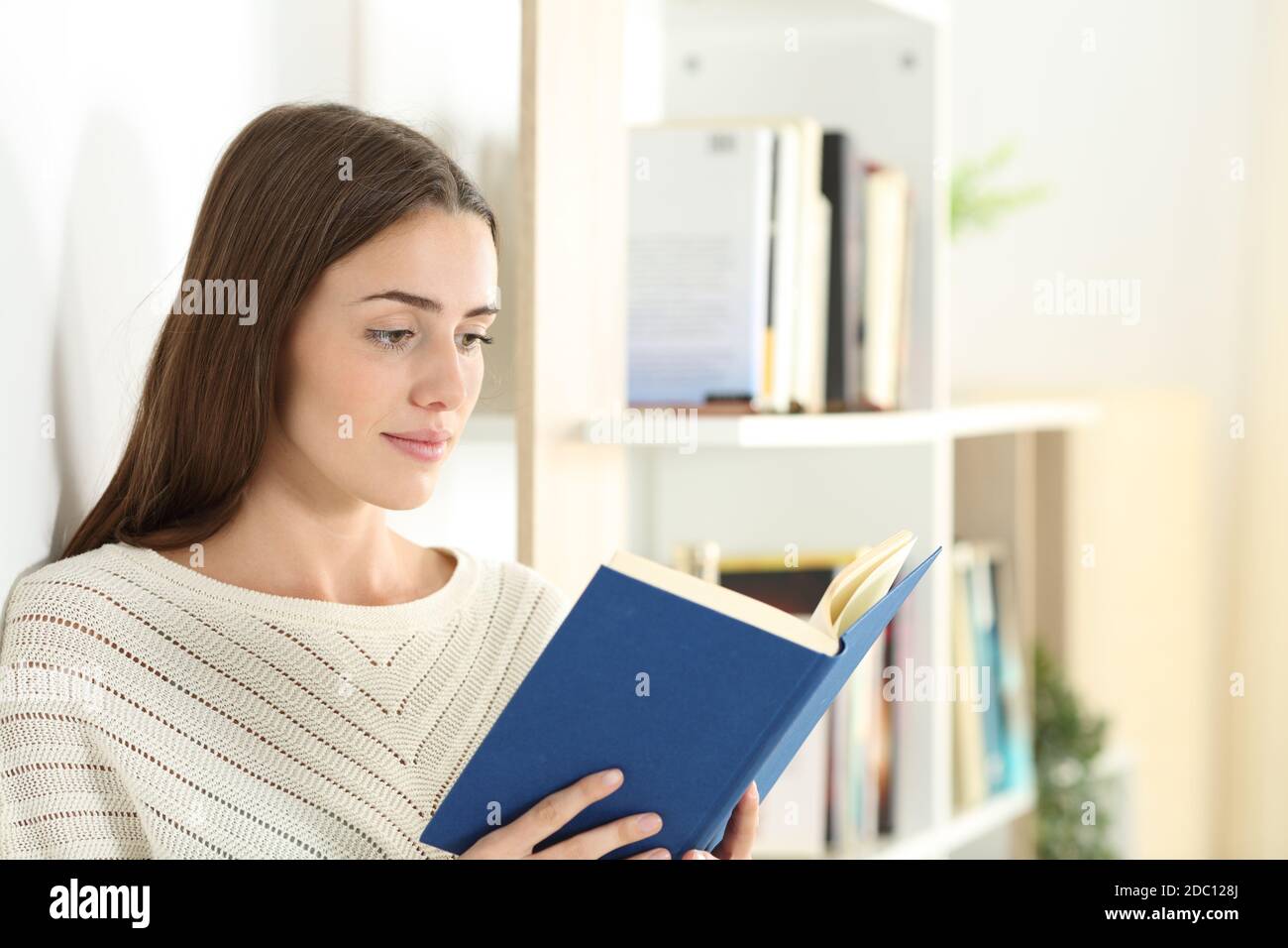 Serious teen reading a paper book standing in the living room at home ...