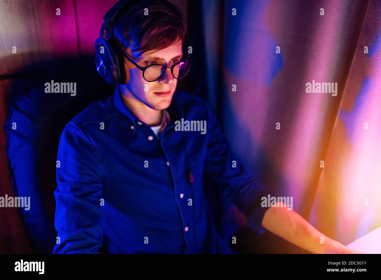 A happy young gamer with glasses and a headset plays computer games at ...