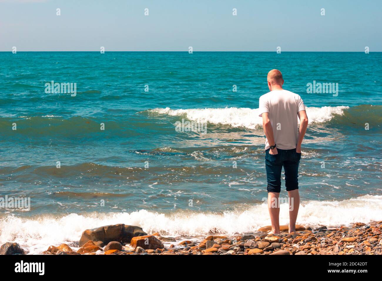 young man stands on the seashore with waves and looks at the water ...