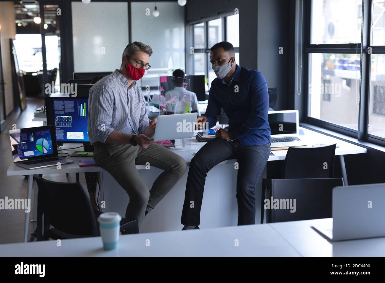 Diverse male colleagues wearing face masks using laptop while sitting ...