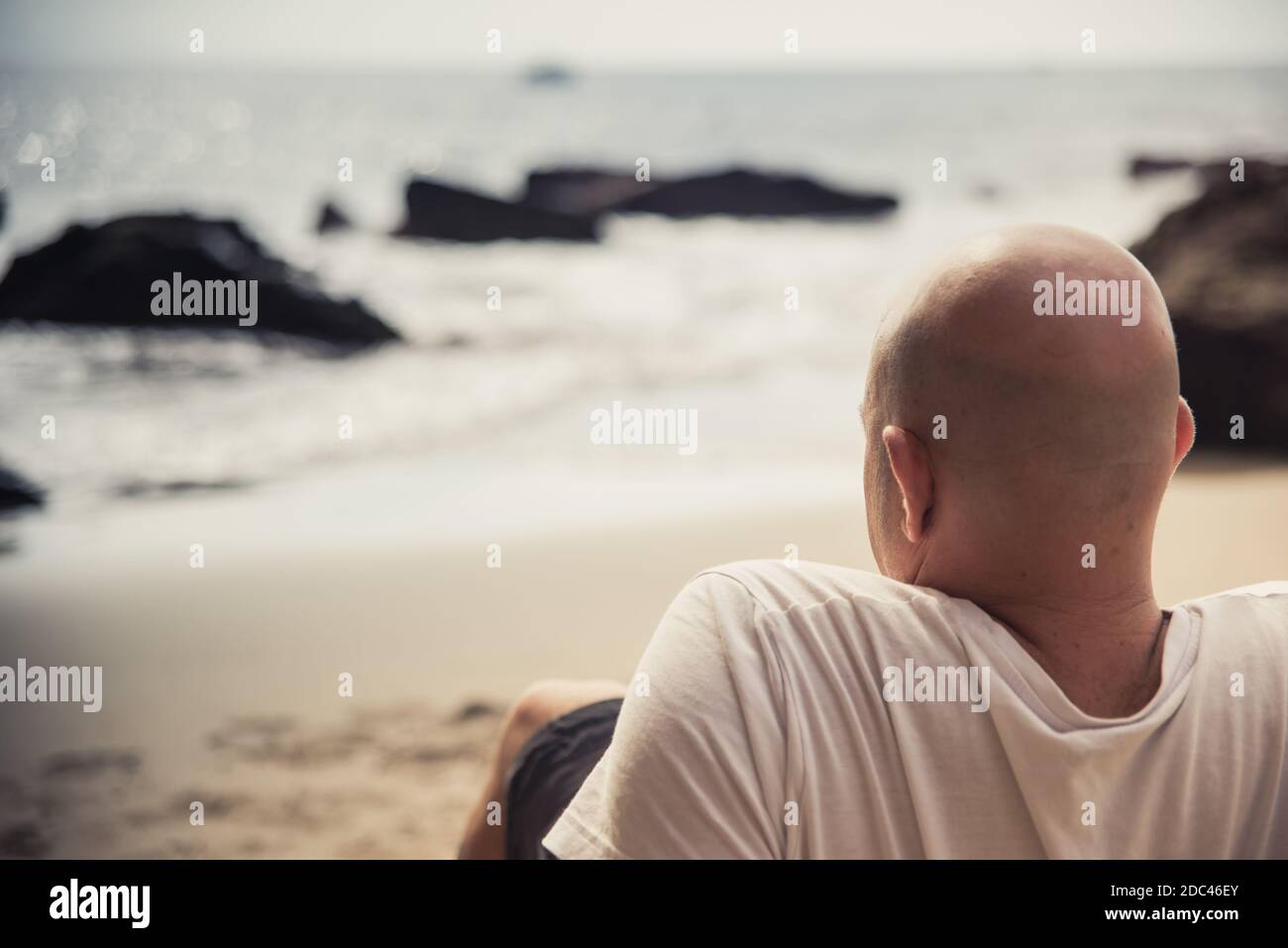 bald adult man on his back sitting and relaxed on the sand looking out ...