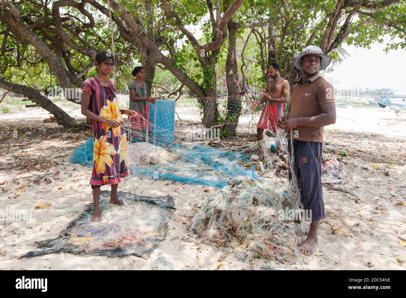 Fishermen and women repair their fishing nets under the shade of a tree ...