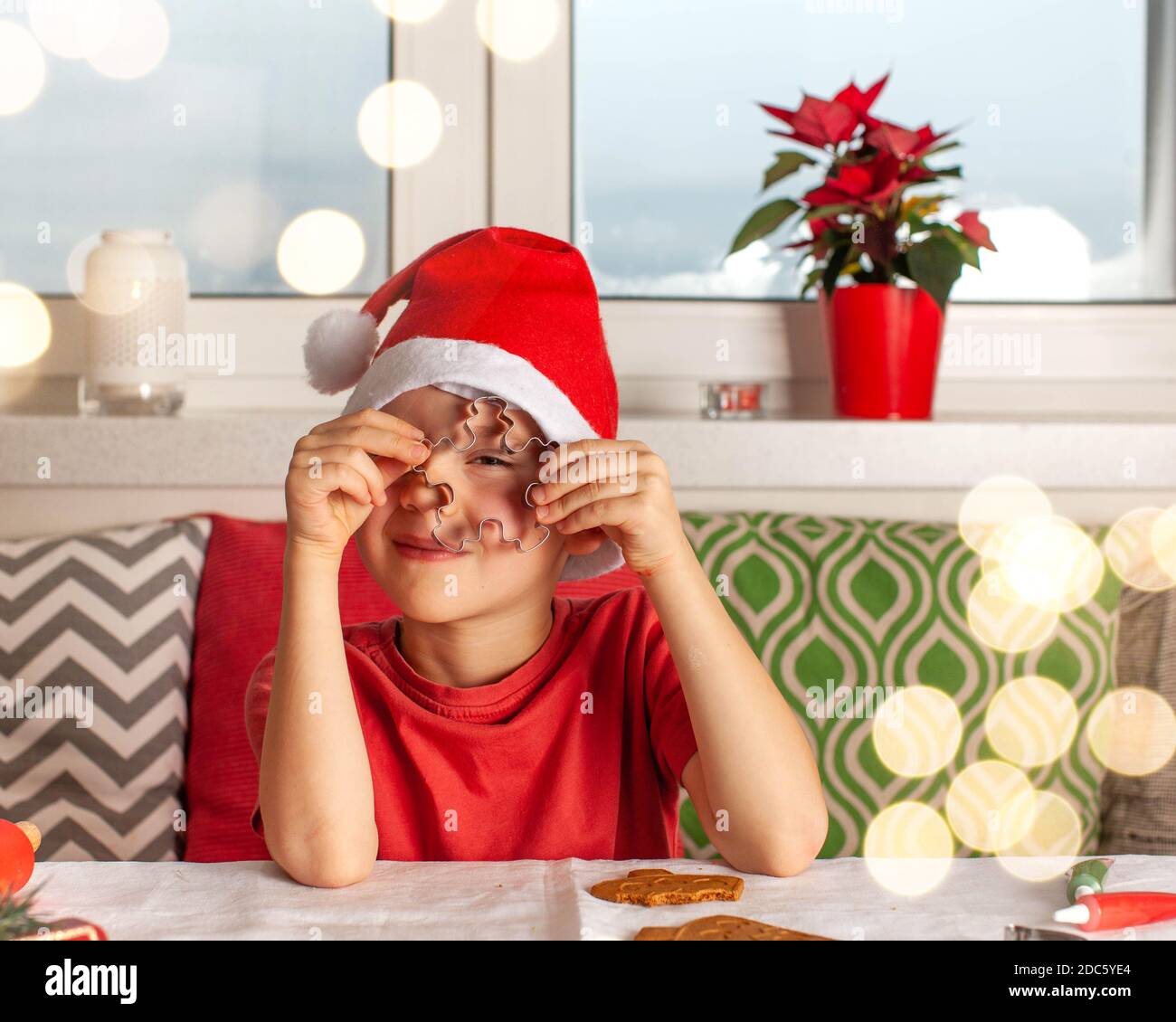 Boy in a Santa Claus hat looks playfully through a cookie cutter ...