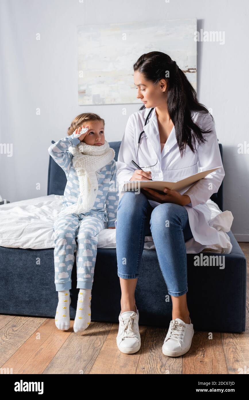 doctor writing in outpatient card while sitting near diseased girl ...