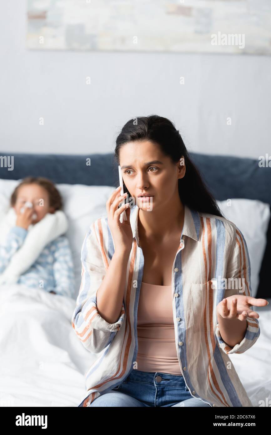 worried woman talking on mobile phone near sick daughter lying in bed ...