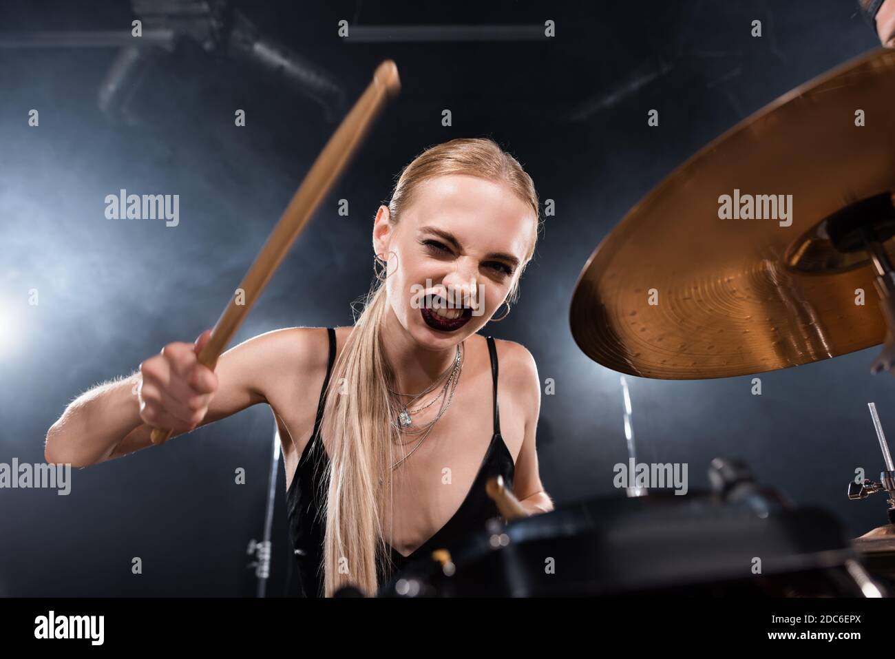 Blonde woman with drumstick winking and playing drum plate on blurred ...